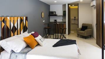 Quarto com cama, almofadas e mesa de cabeceira. Inclui uma kitchenette equipada com micro-ondas e uma cadeira.