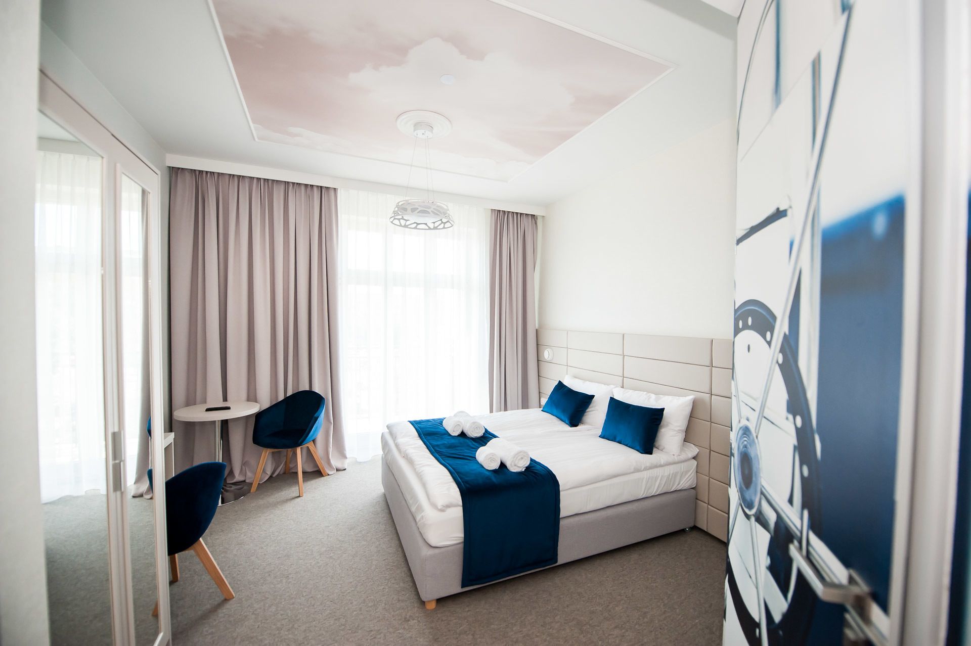 Quarto de hotel com cama, escrivaninha, cadeira e uma grande janela com cortinas.