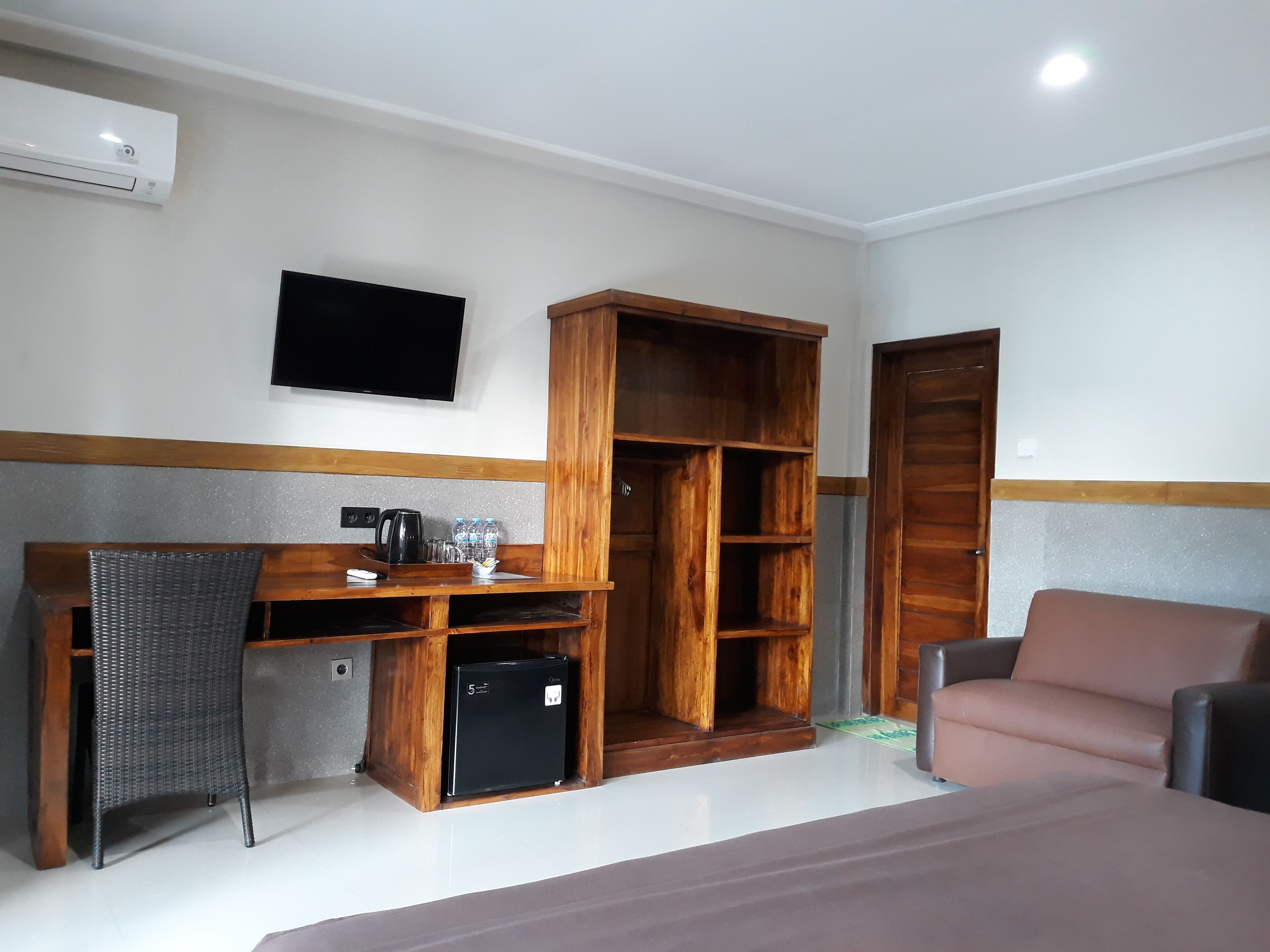Quarto de hotel com uma cama, uma escrivaninha com computador, um pequeno frigorífico, guarda-roupa de madeira, televisão e uma porta.