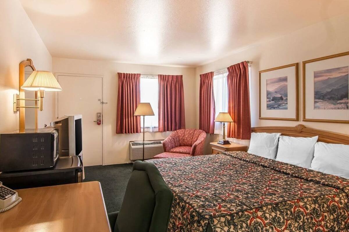 Quarto de hotel com cama, escrivaninha com computador, cadeira, abajur e janela com cortinas.