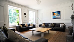 Uma sala de estar ampla com dois sofás de couro preto, uma mesa de centro de madeira e grandes janelas que permitem entrada de luz natural.