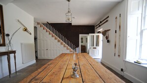 Uma mesa de madeira com copos e um vaso, uma escadaria e uma porta que dá para outro quarto.