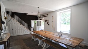 Quarto espaçoso com uma grande mesa de madeira, escada com corrimão branco e janela com vista para o verde.