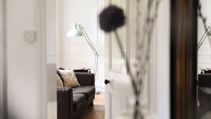 Uma porta dá acesso a um quarto com um sofá de couro escuro, uma luminária branca e um vaso com uma única flor.