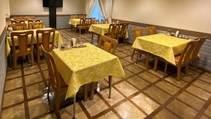 Uma sala de jantar com várias mesas e cadeiras, uma cruz na parede e uma janela com cortinas.
