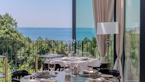 Mesa de jantar posta com taças de vinho, pratos e talheres, com vista panorâmica para o mar e vegetação.