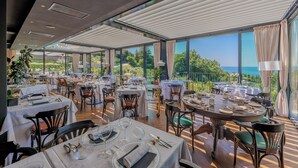 Um restaurante com mesas preparadas para refeições, amplas janelas com vista para o mar e um teto com design moderno.