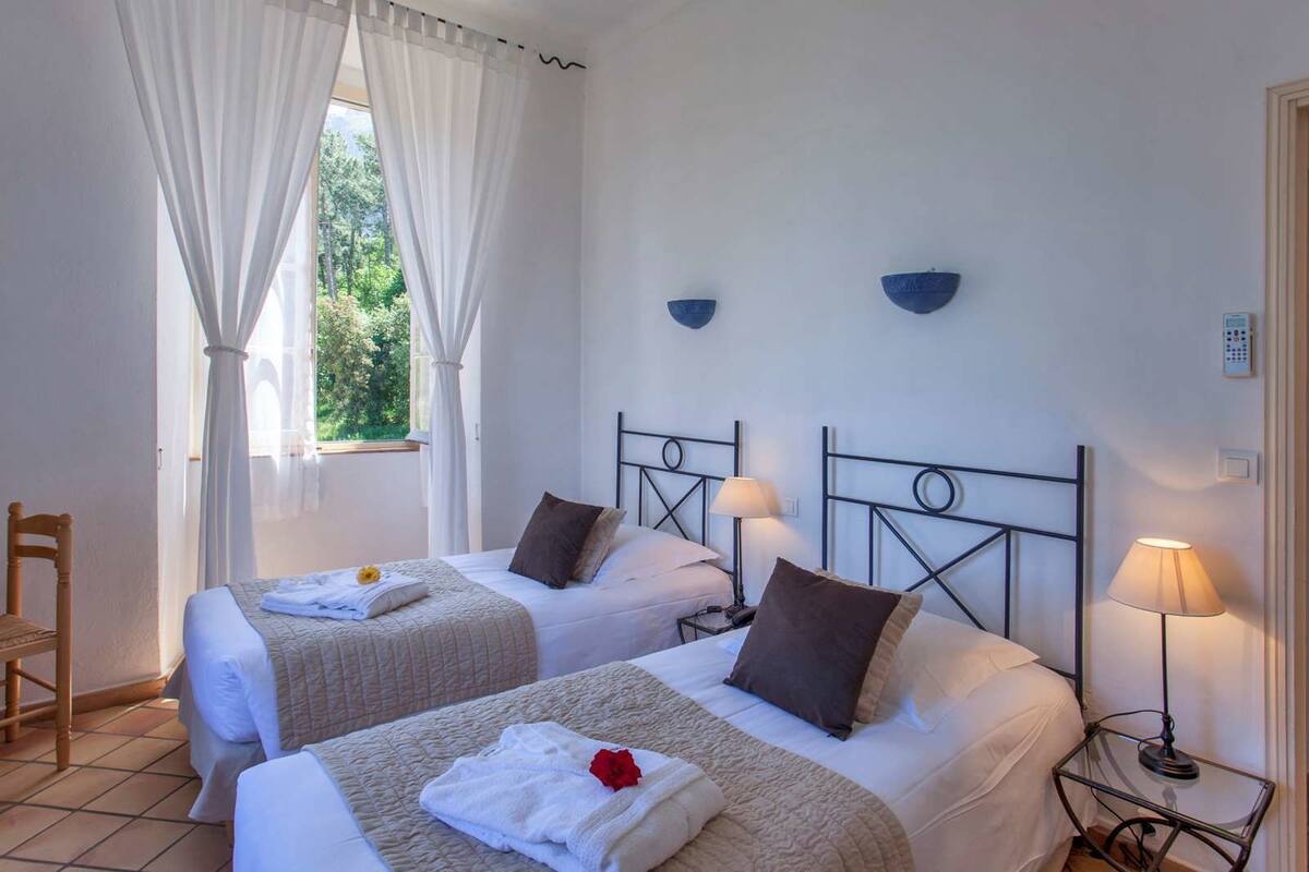 Quarto de hotel com duas camas, uma cadeira, uma janela com cortinas e uma mesa de cabeceira com abajur.