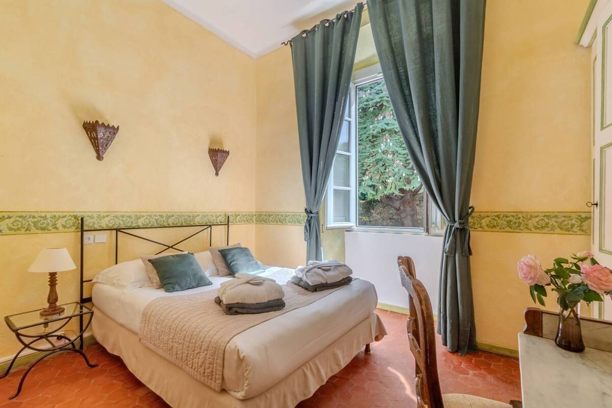 Quarto com cama, mesa de cabeceira, abajur e janela com cortinas.