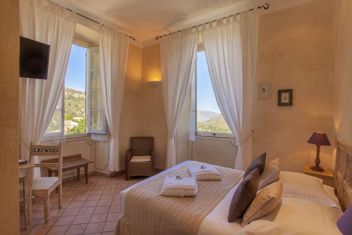 Quarto com cama, cadeira, escrivaninha, televisão e janela com cortinas.