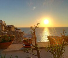 Vista costeira com pôr do sol sobre o mar, edifícios ao fundo e plantas em vasos no primeiro plano.