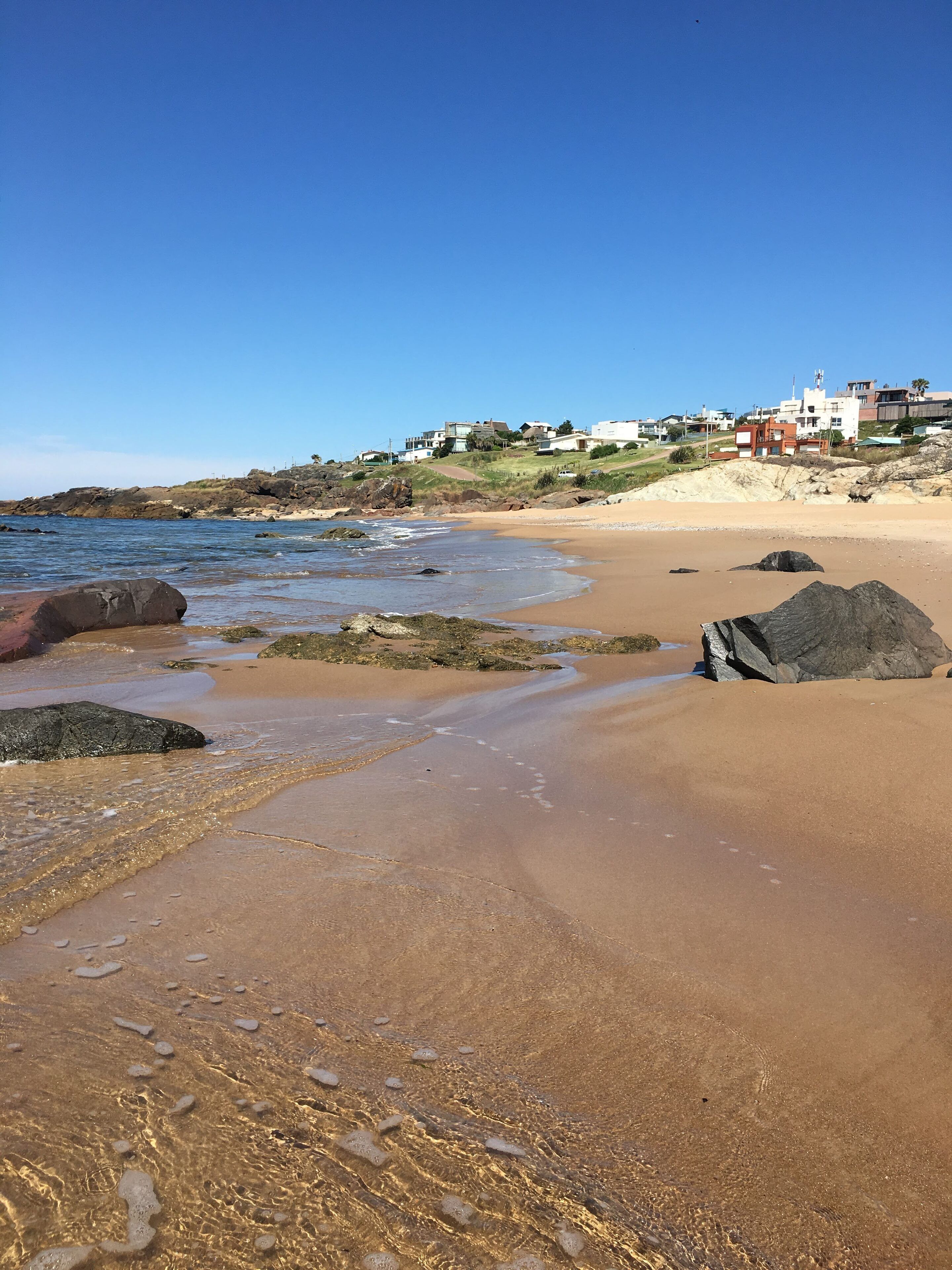 Uma praia com águas cristalinas, rochas e casas ao fundo.