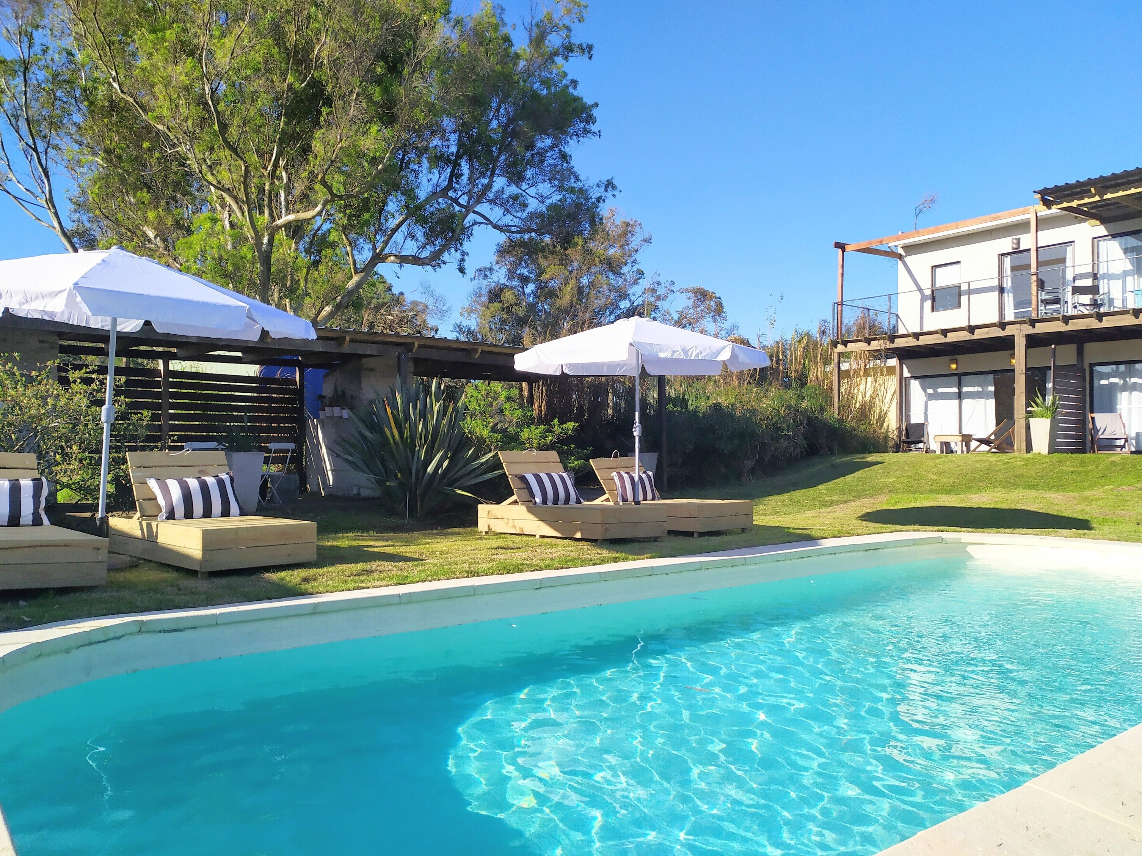 Piscina com espreguiçadeiras e guarda-sóis, cercada por vegetação e um edifício.