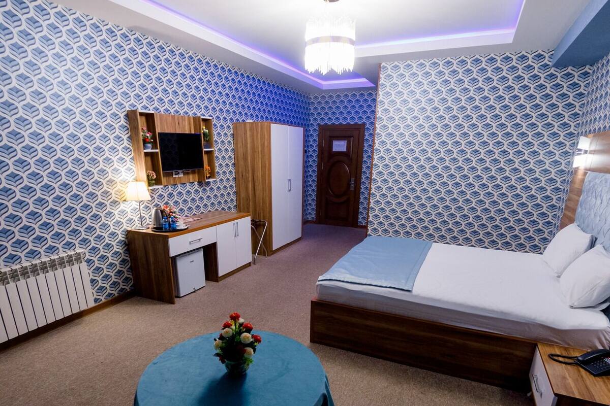 Quarto de hotel com papel de parede azul e branco estampado, uma mesa redonda com um vaso de flores, uma cama com colcha azul, uma escrivaninha com uma luminária e uma televisão na parede.