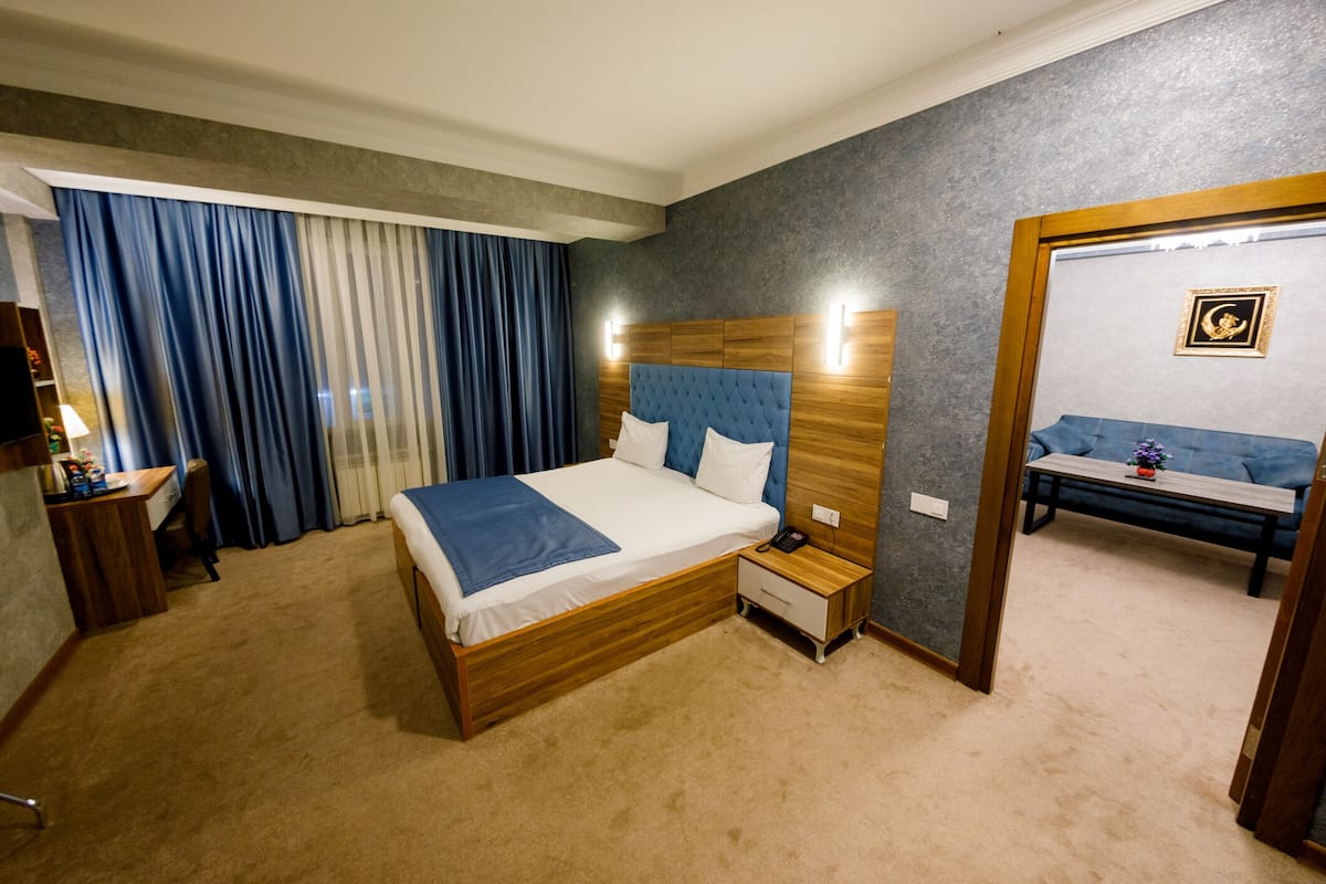 Quarto de hotel com cama de madeira, mesa de cabeceira, escrivaninha com cadeira, sofá azul e uma grande janela com cortinas.