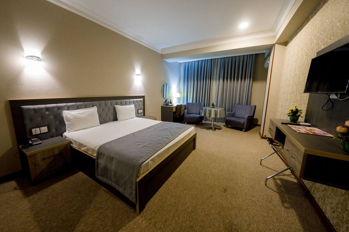 Quarto de hotel com uma cama grande, mesas de cabeceira, escrivaninha, cadeira, televisão e janela com cortinas.
