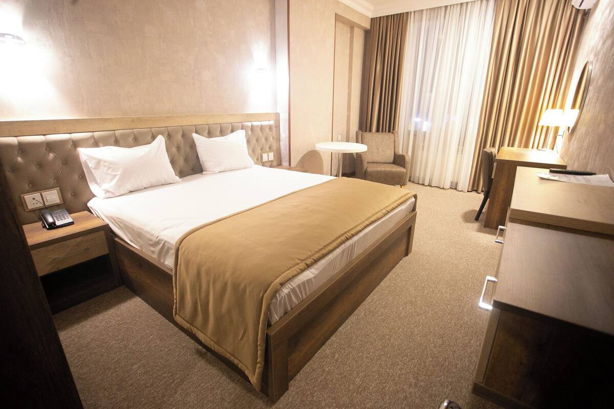 Quarto de hotel com uma cama grande, uma mesa de cabeceira com telefone, uma pequena mesa redonda, uma cadeira e uma escrivaninha com abajur.