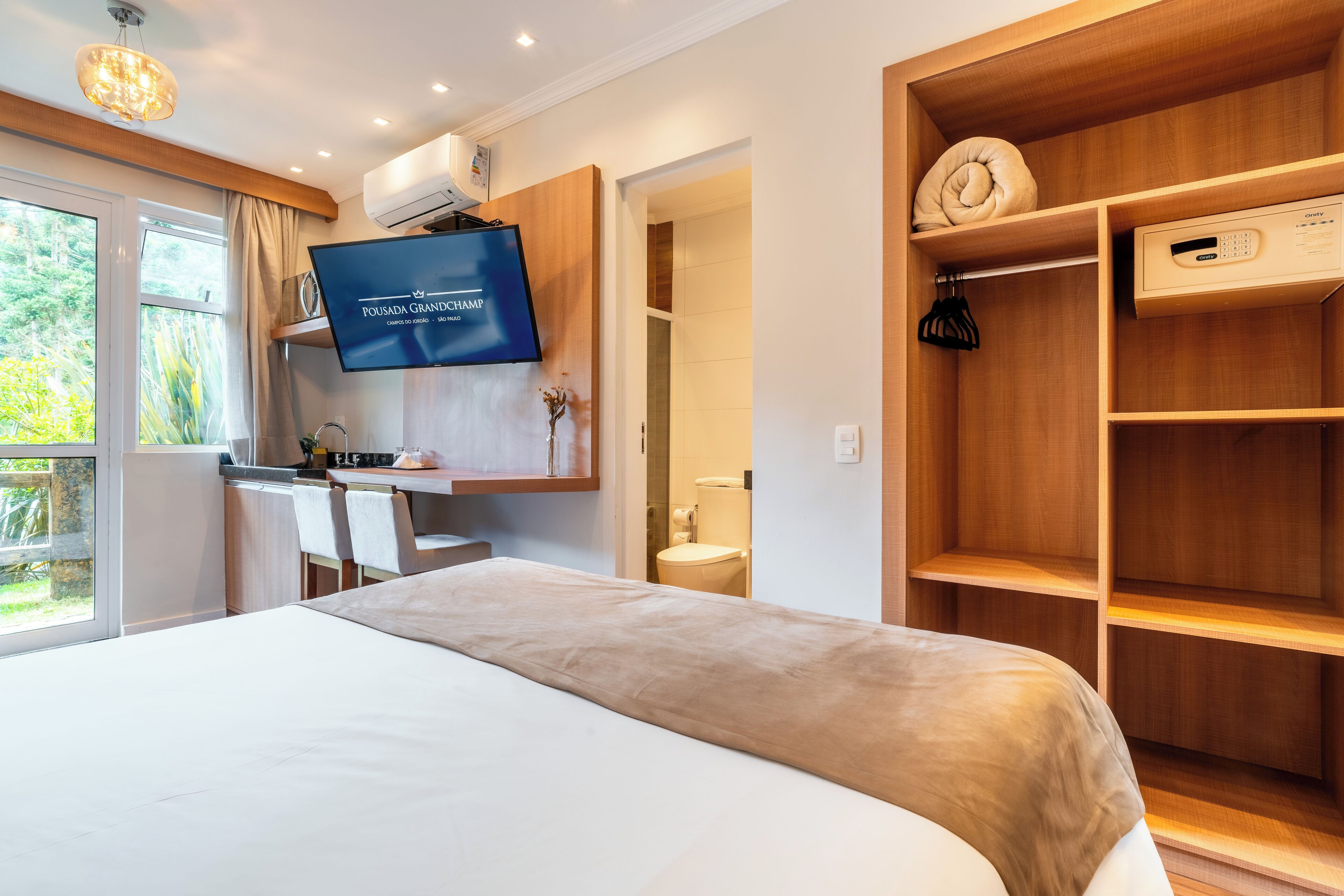 Quarto de hotel com cama, televisão fixada na parede, guarda-roupa de madeira e secretária com cadeira.