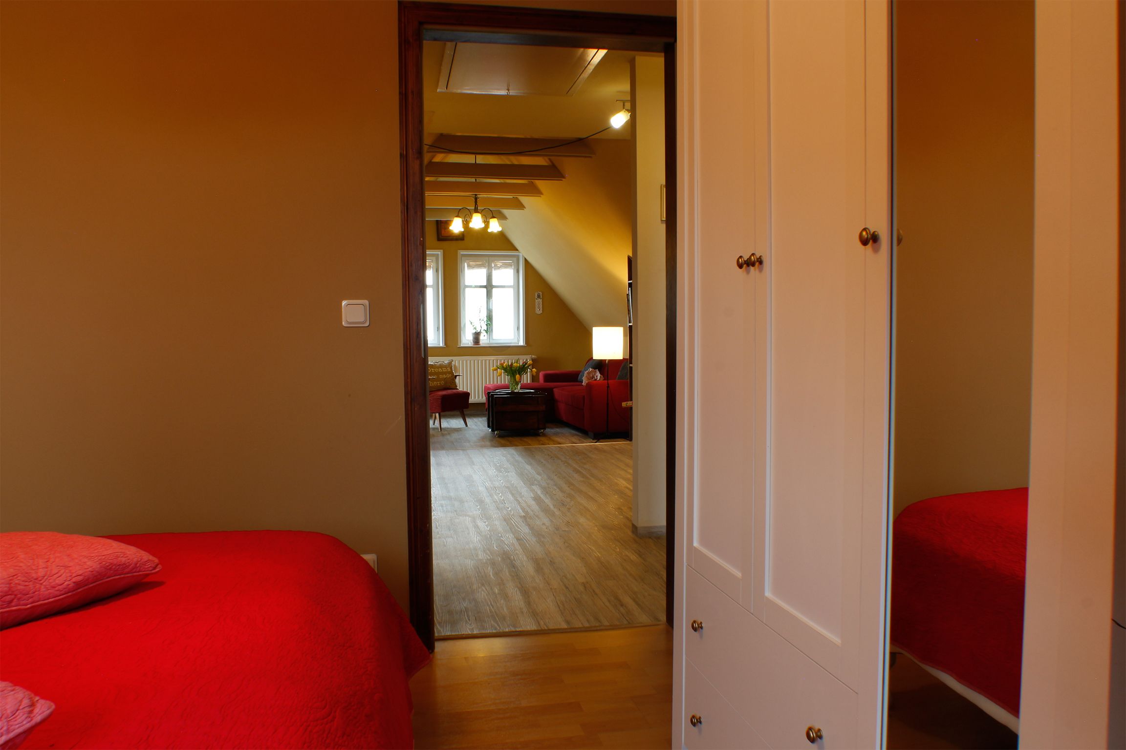 Quarto com piso de madeira, cama vermelha e sofá vermelho. Há uma porta que dá para outro quarto.