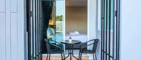 Pequena varanda com mesa e duas cadeiras, porta de correr de vidro e vista para um quarto com cama e janela.