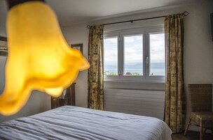 Quarto com cama, janela com cortinas, uma cadeira e vista para o mar.