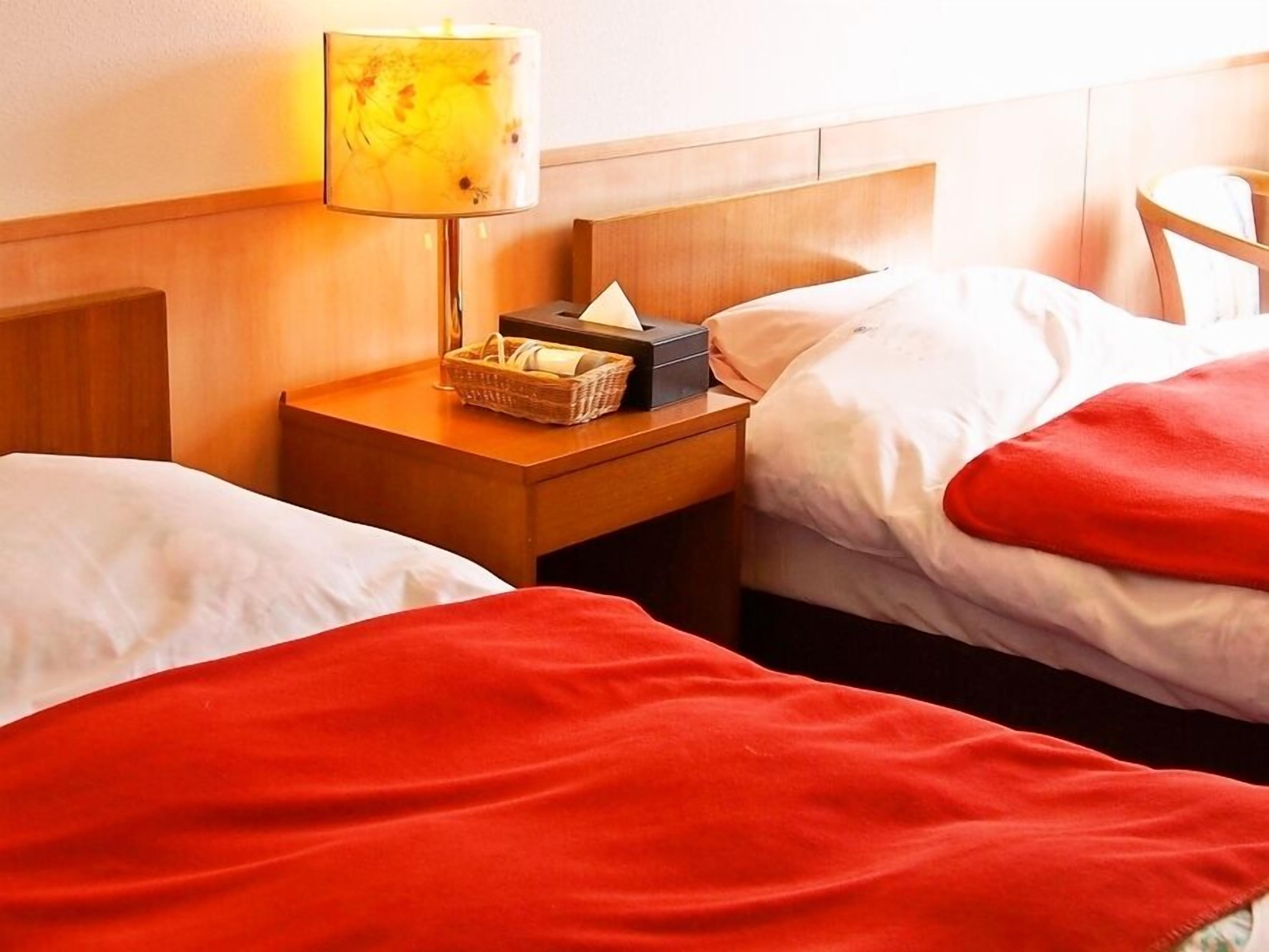 Quarto de hotel com duas camas, uma mesinha de cabeceira com abajur e uma cesta.
