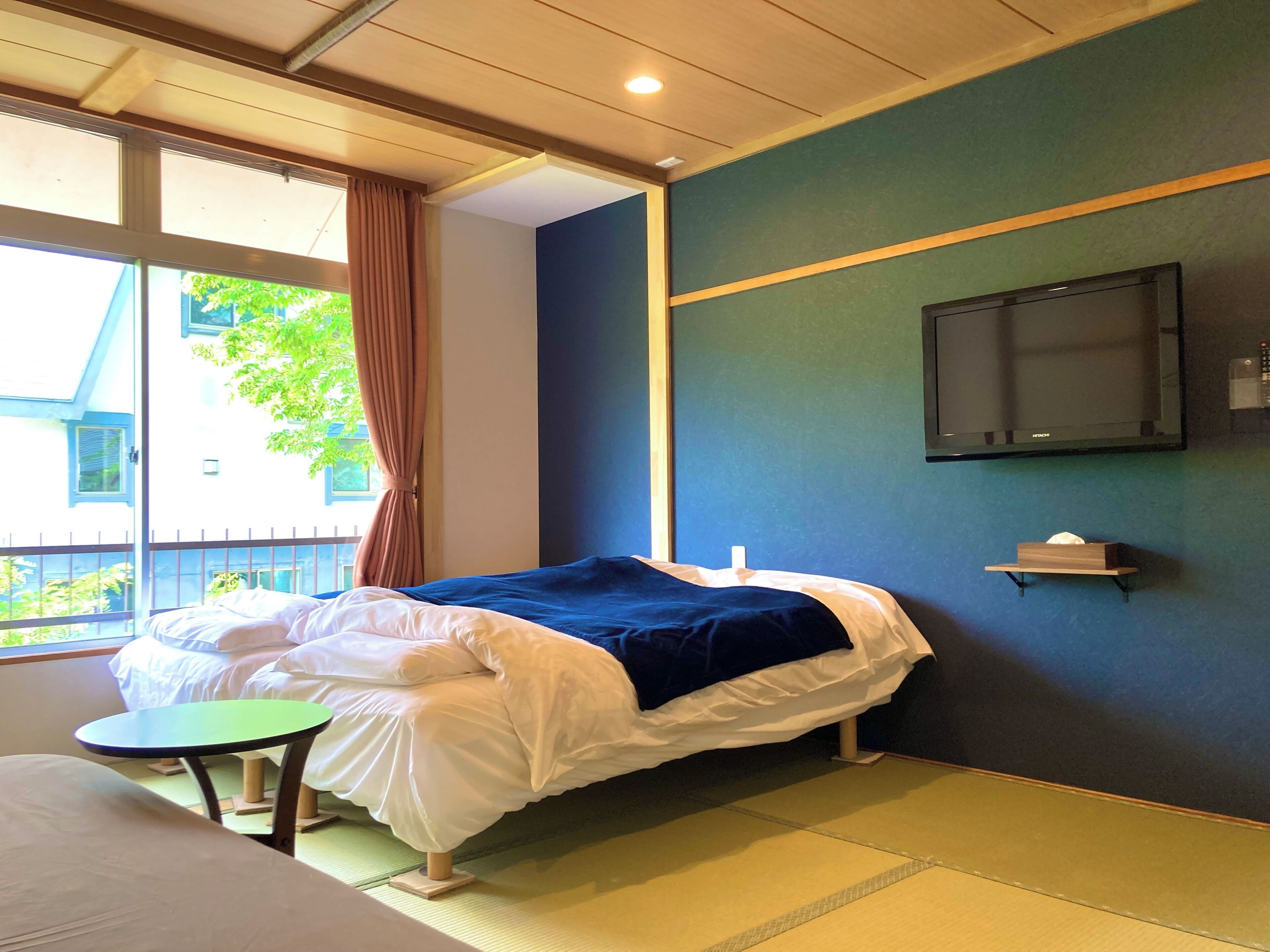 Quarto de hotel com cama, uma mesinha, televisão fixada na parede e janela com cortinas.