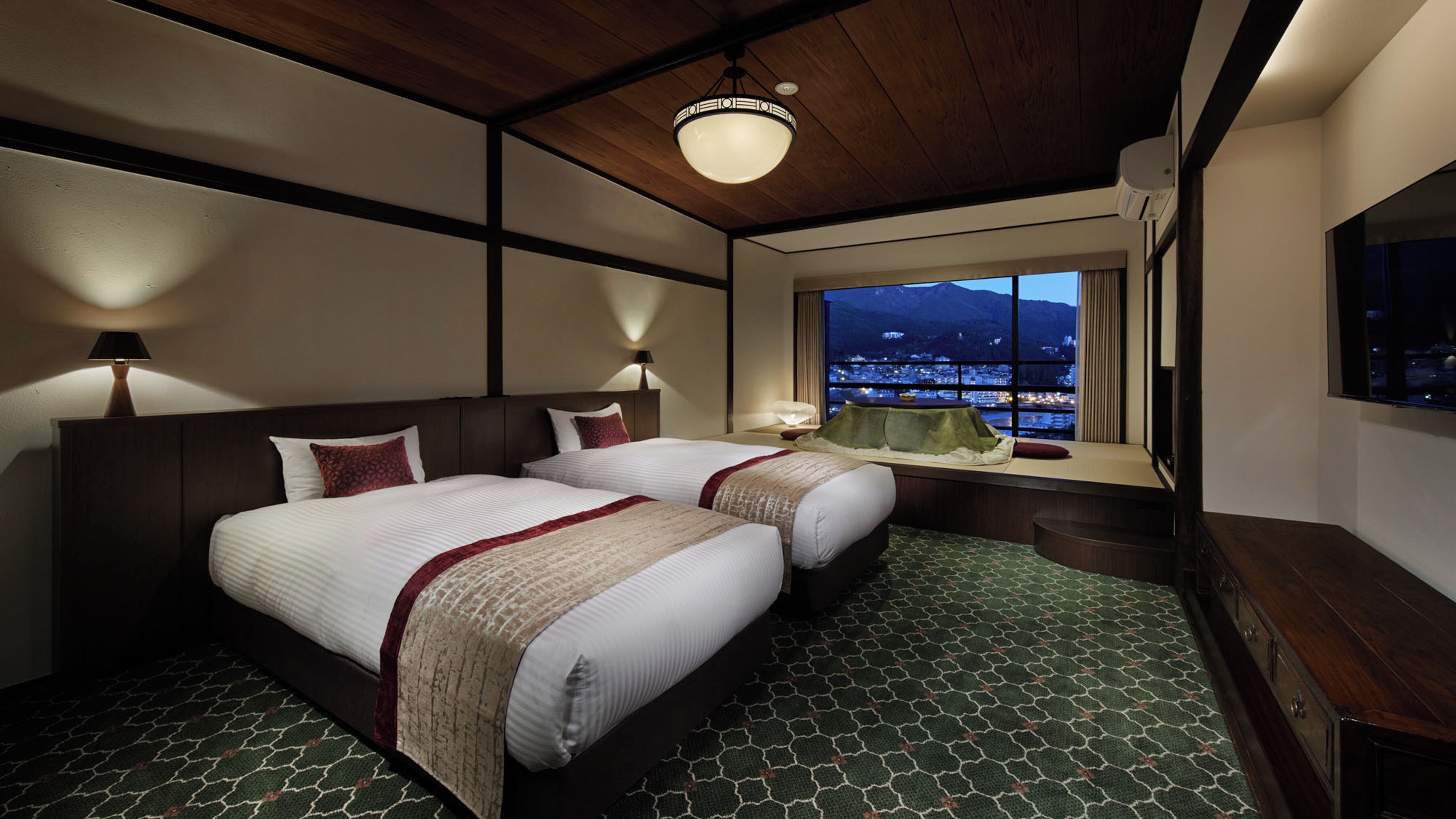 Quarto de hotel com duas camas, uma janela grande com vista para a cidade, teto de madeira e um tapete verde estampado.