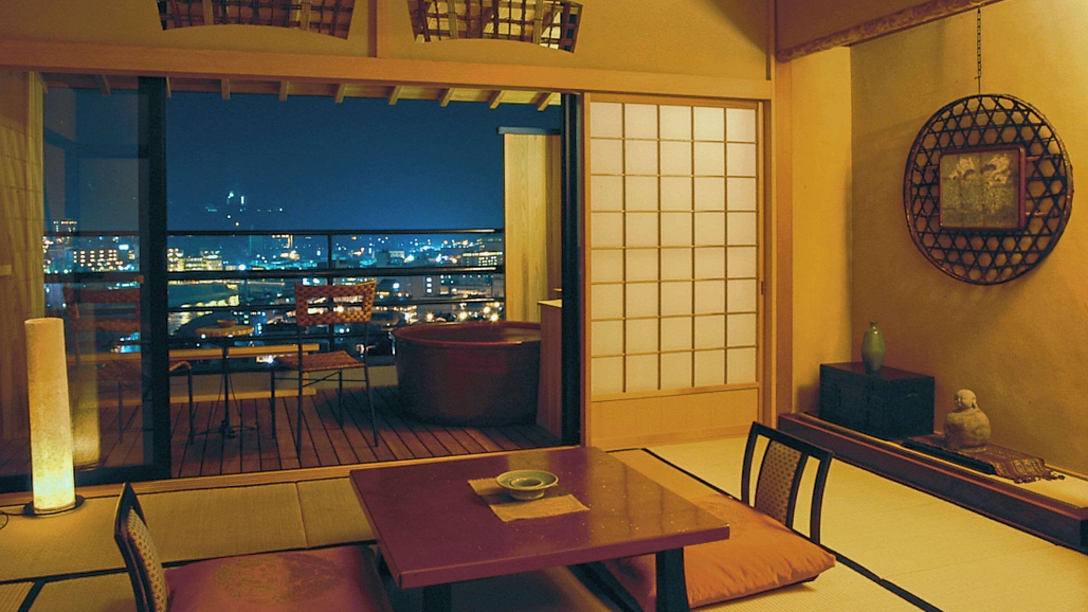 Quarto com vista para a paisagem urbana à noite, porta de correr de vidro, mesa com almofada e um biombô japonês tradicional.