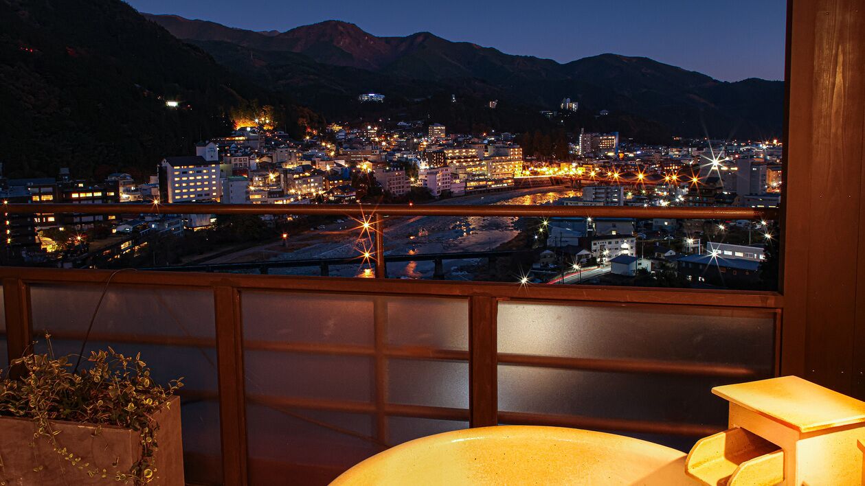Vista noturna de uma cidade com um rio, montanhas ao fundo e uma varanda com vista.