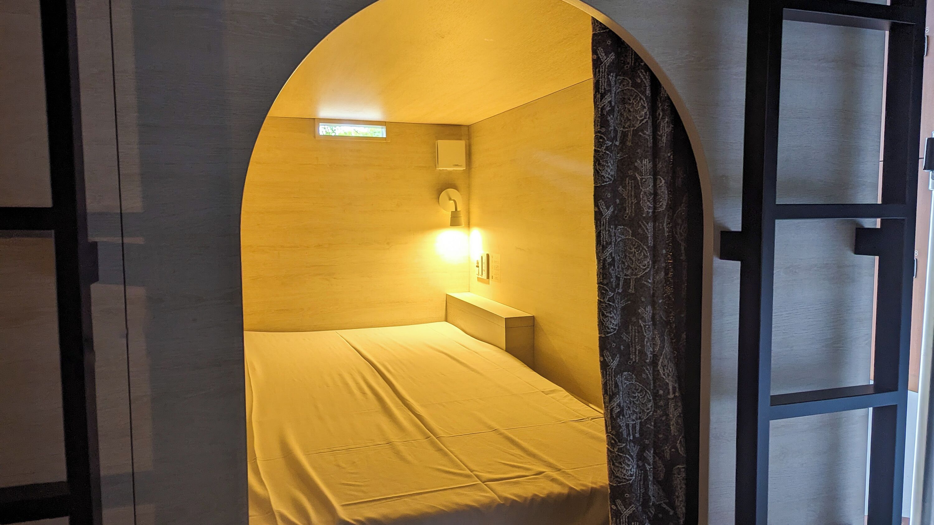 Uma cama de solteiro num quarto com parede curva, janela pequena e luminária fixada na parede.
