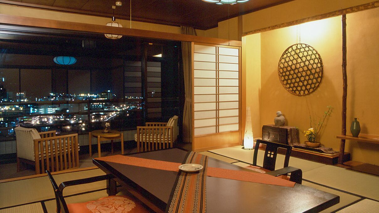 Um quarto japonês tradicional com tatames, uma mesa baixa e vista para a cidade à noite.