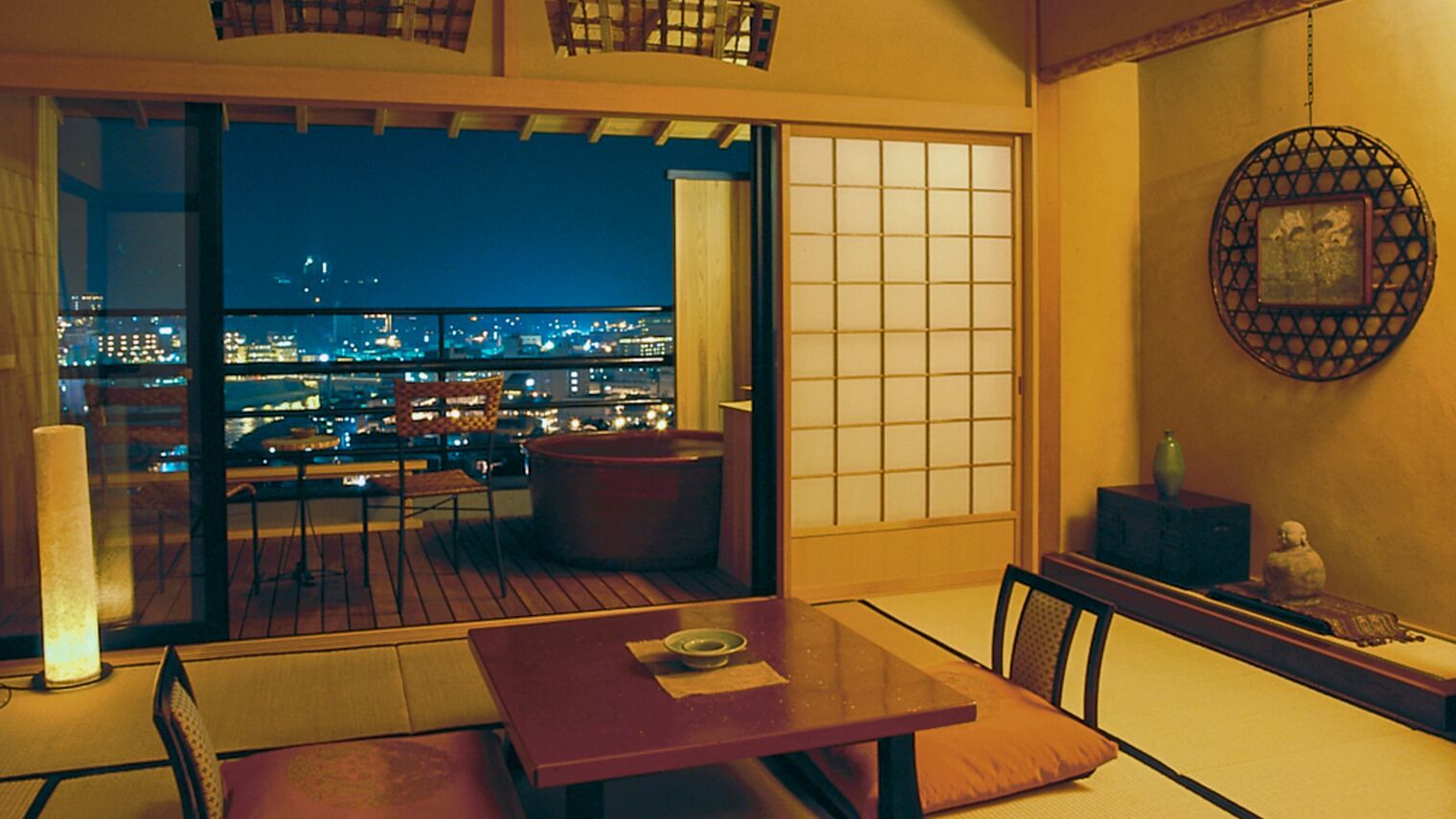Quarto com vista para a cidade à noite, porta deslizante, mesa com almofada e objeto decorativo na parede.