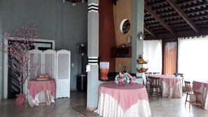 Uma sala de jantar com mesas cobertas por toalhas xadrez vermelho e branco, uma planta florida rosa decorativa e um teto rústico.