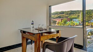 Quarto com mesa de vidro, uma cadeira, uma garrafa de água e janela com vista para uma paisagem verde e edifícios.