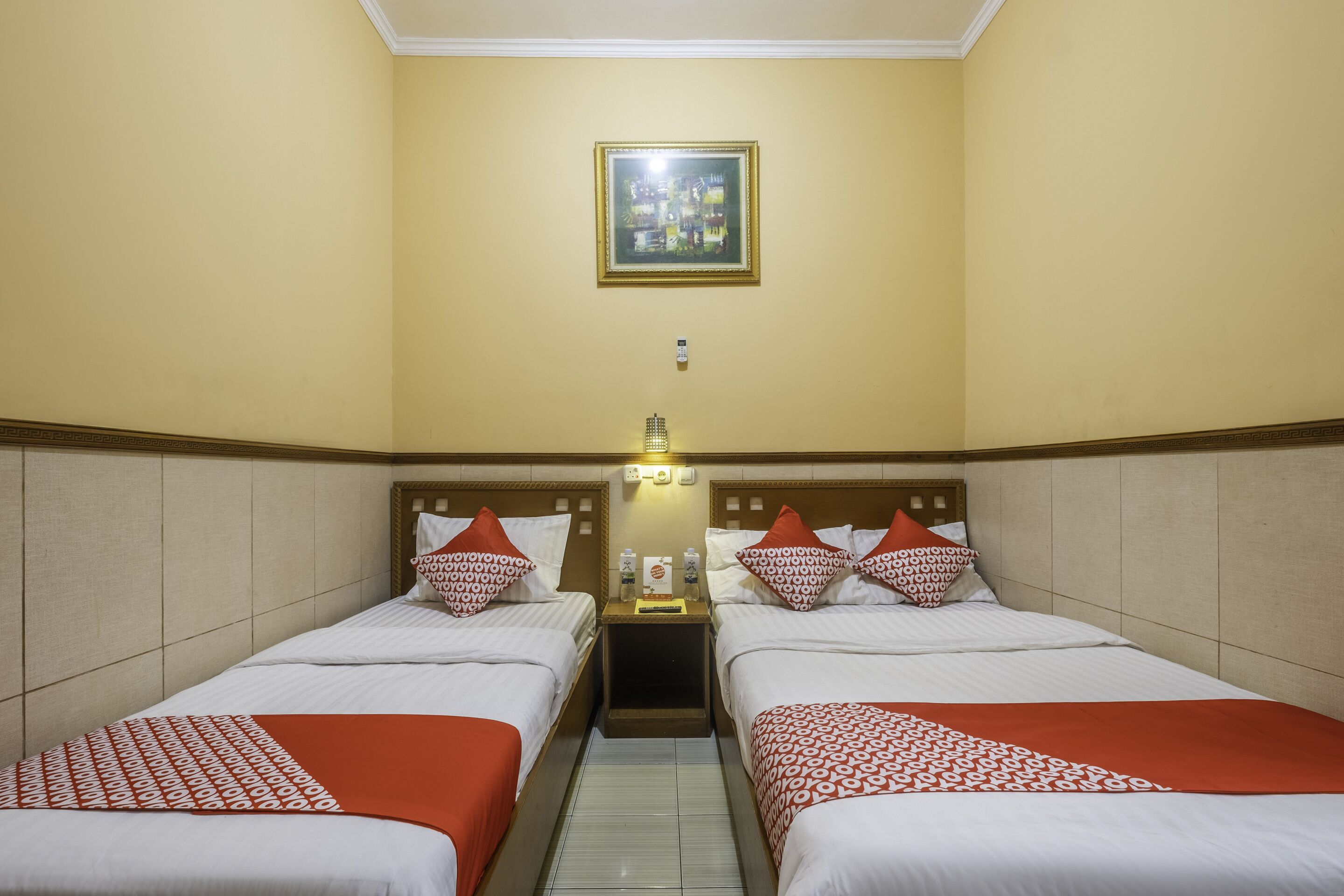 Quarto de hotel com duas camas, cada uma com roupa de cama vermelha e branca, uma mesa de cabeceira com abajur e um quadro na parede.