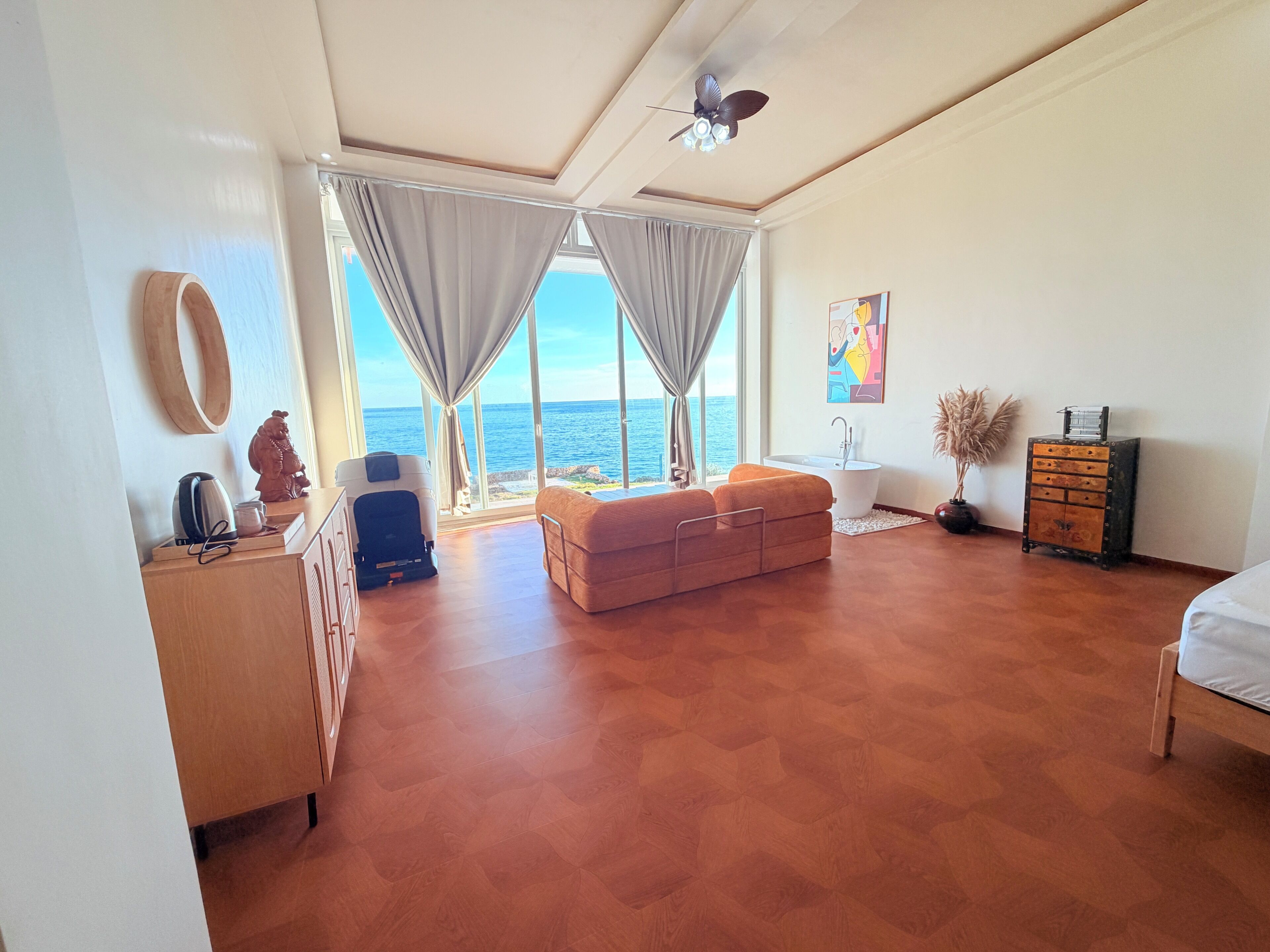 Um quarto espaçoso com uma grande janela, um ventilador de teto, um sofá, uma cama e uma cômoda de madeira.