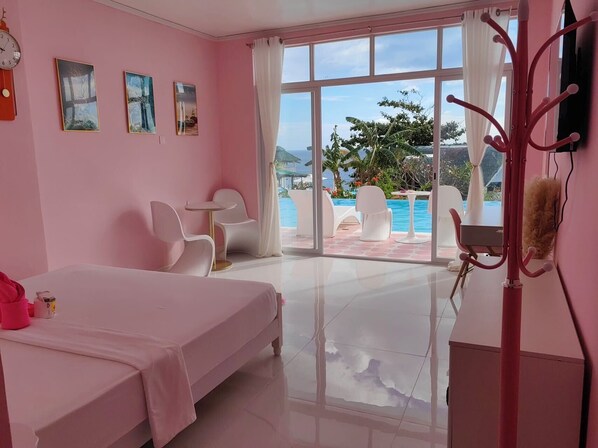 Quarto de hotel em tons de rosa, com cama, duas cadeiras, uma mesinha e vista para a piscina e o oceano.