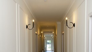 Um corredor comprido e estreito, com paredes brancas e soalho de madeira, com arandelas nas paredes e um radiador à esquerda.