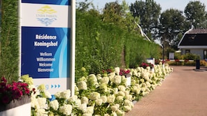 Placa de UNIEK PARKEN com mensagem de boas-vindas em vários idiomas, rodeada por um caminho com flores brancas e vegetação.