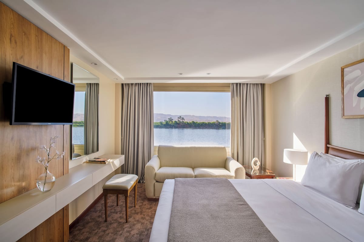 Quarto de hotel com uma cama grande, televisão na parede, sofá, escrivaninha e vista para um lago.
