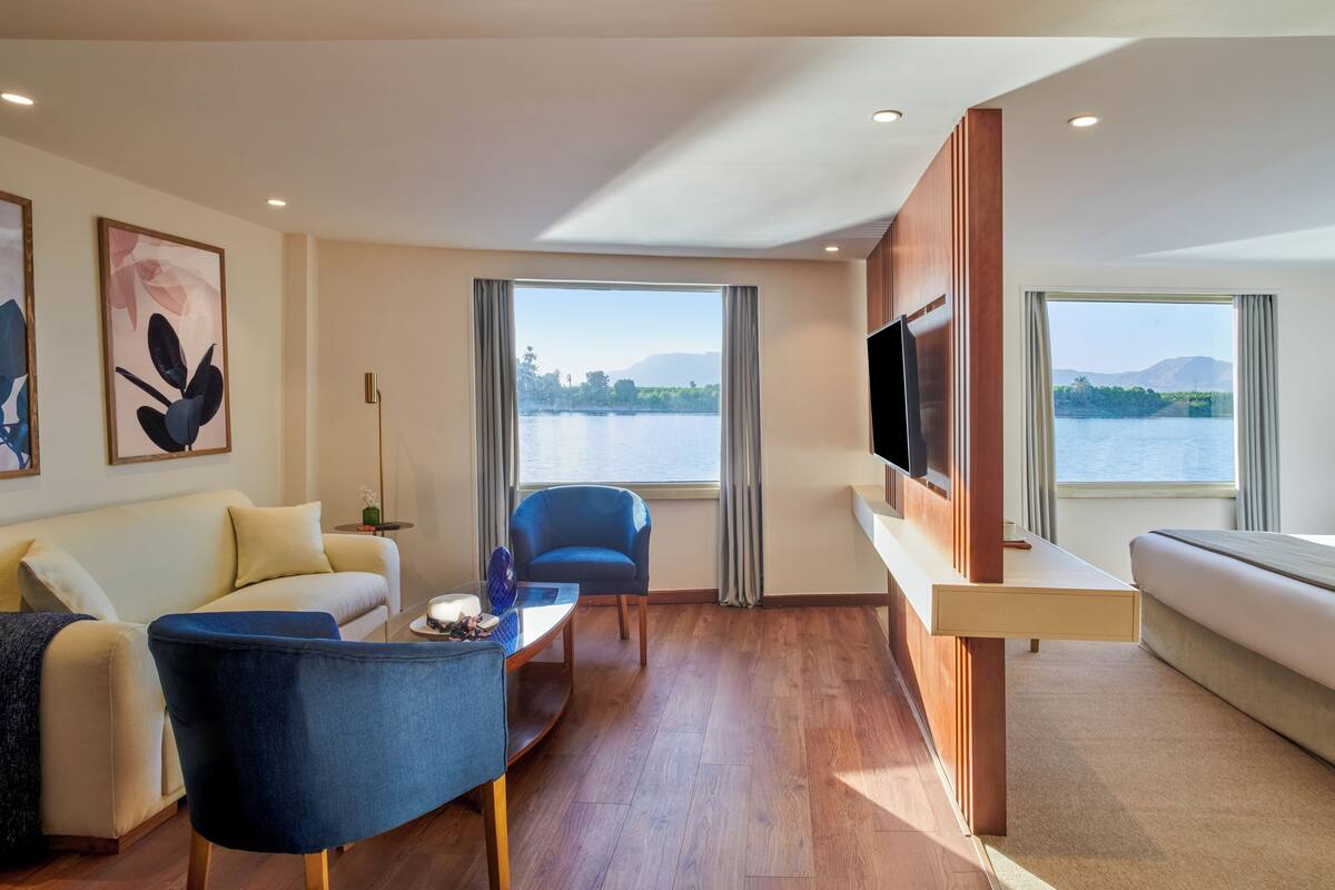 Uma sala de estar moderna com um sofá, uma poltrona azul, uma mesa de centro e uma TV na parede. Janelas amplas oferecem vista para um lago e montanhas.