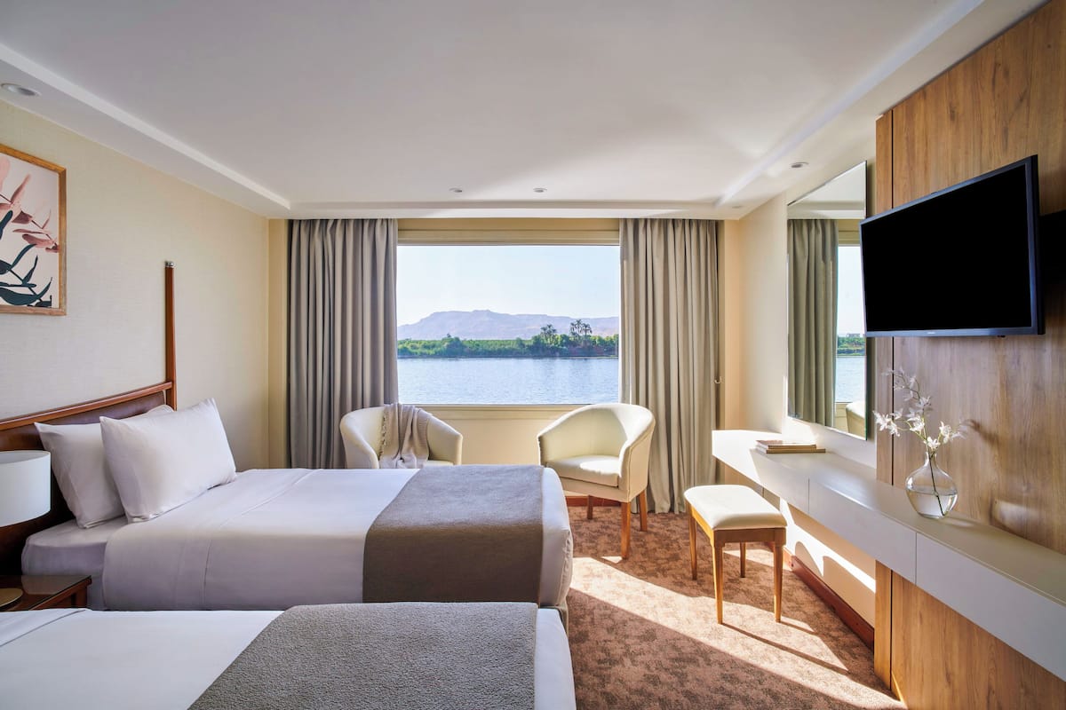 Quarto de hotel com duas camas, uma mesa, uma cadeira, televisão e vista para um corpo de água.