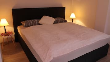 Uma cama bem arrumada com um cobertor branco, duas almofadas e uma cabeceira escura. Há duas luminárias de cabeceira de cada lado da cama.