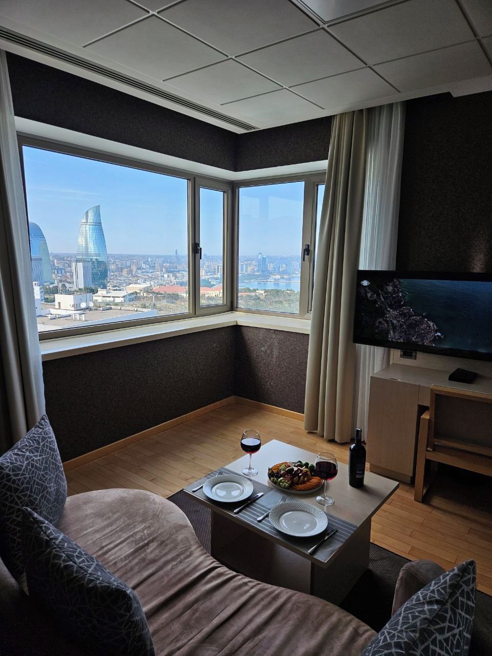 Quarto de hotel moderno com uma janela grande que oferece vista para a cidade, uma mesa de jantar posta para dois e uma TV de tela plana em um suporte.