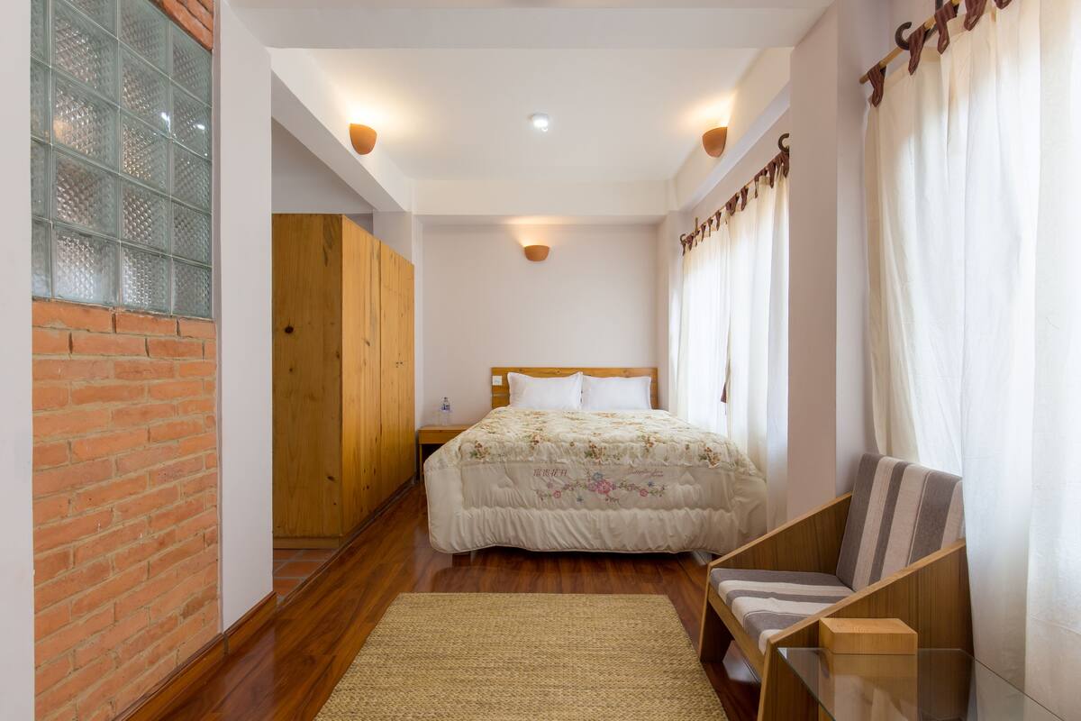 Quarto com cama de madeira, mesa de cabeceira, cadeira, parede de tijolos e janela com vista.