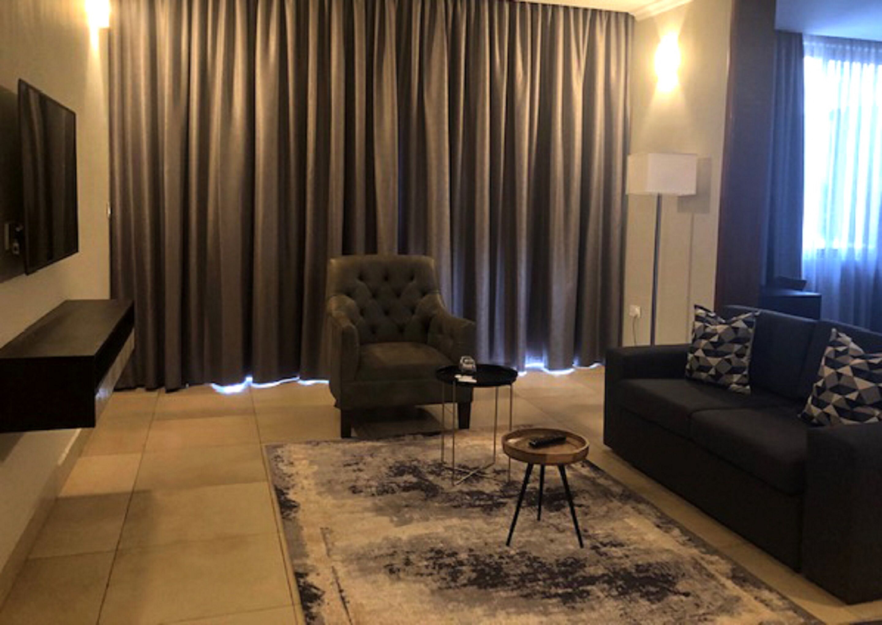 Uma sala de estar com poltrona escura, sofá com almofadas estampadas, mesa de centro e uma grande janela com cortinas.