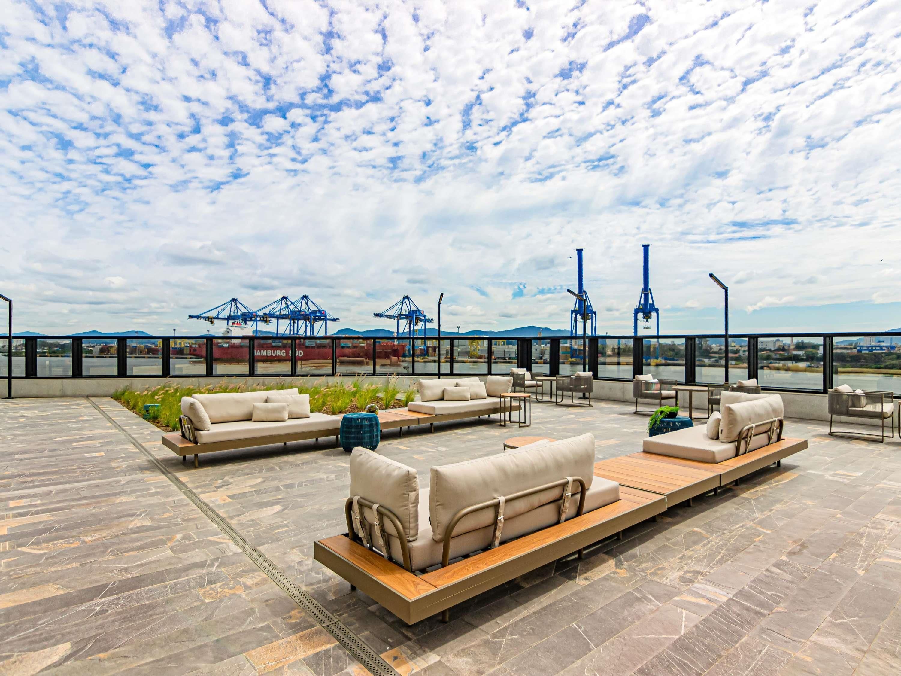 Um terraço com área de estar ao ar livre, vista para o porto e guindastes ao fundo.