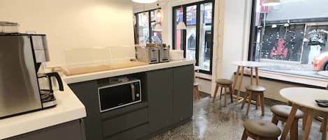 Cozinha moderna com máquina de café, micro-ondas e duas luminárias pendentes. Há banquetas e uma mesa perto de uma janela com vista para a rua.