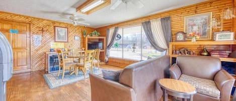Quarto aconchegante com decoração em madeira, ventilador de teto, televisão, sofá, mesa pequena e janela com cortinas.